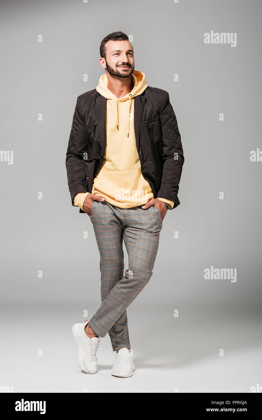 happy stylish man in autumn outfit posing with hands in pockets on grey ...