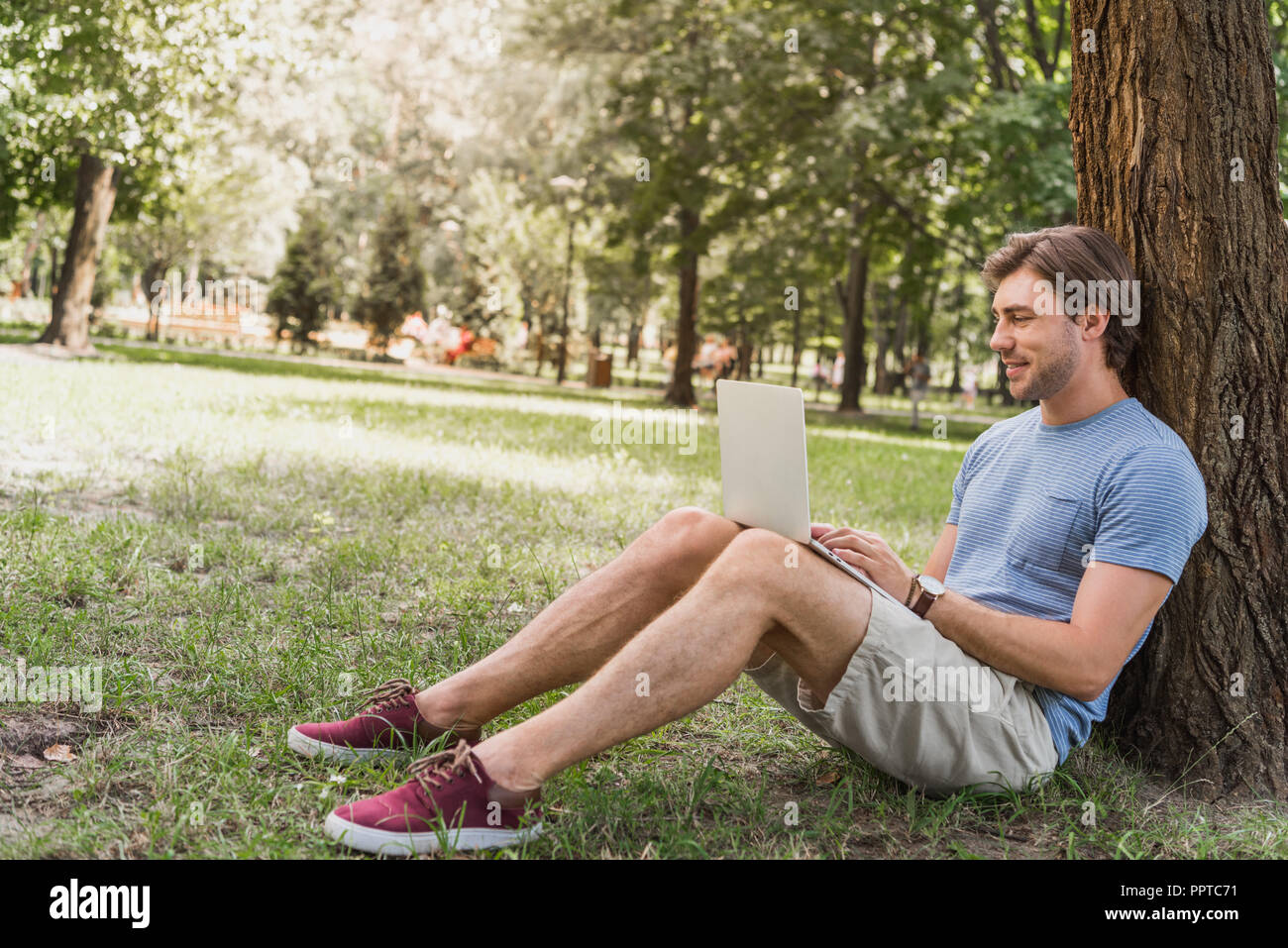 side view of handsome man sitting near tree in park and using laptop ...