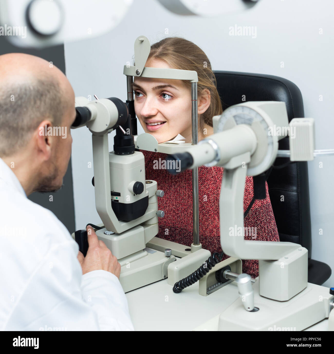 Mature optician doing eye examination of girl with aid of slit lamp ...