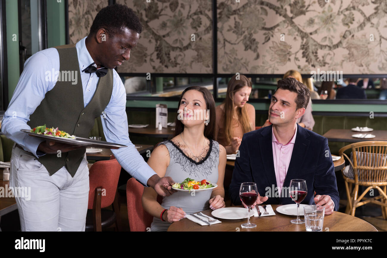 Polite African American waiter bringing ordered dishes to smiling ...