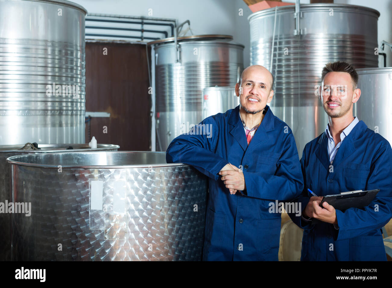 Two smiling male winery employees in uniform working and taking notes ...