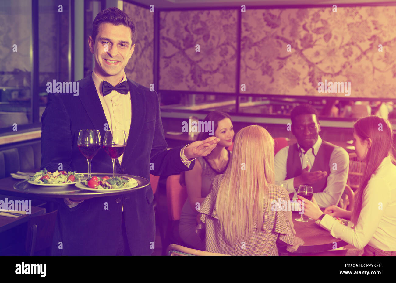 Polite waiter holding tray at restaurant with customers behind him ...