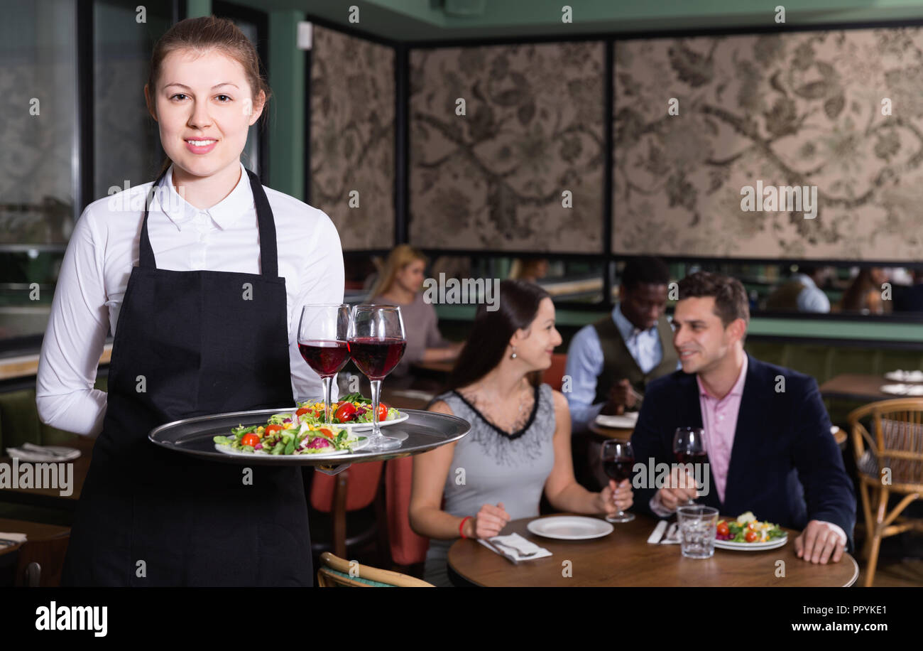 Attractive positive smiling young waitress with serving tray welcoming ...