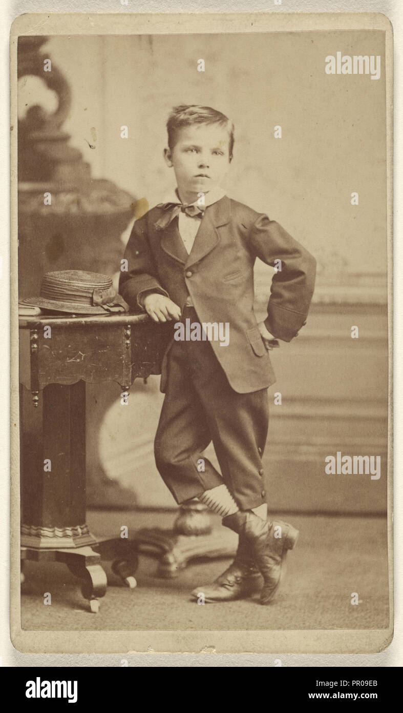 Young boy in suit standing and posed near a table with a hat on top ...