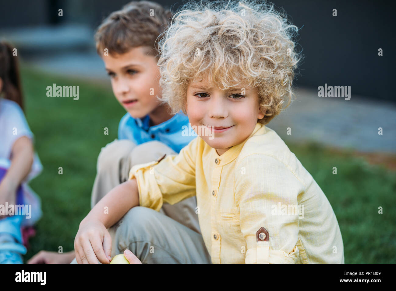 adorable curly schoolboy sitting on grass with classmate Stock Photo ...