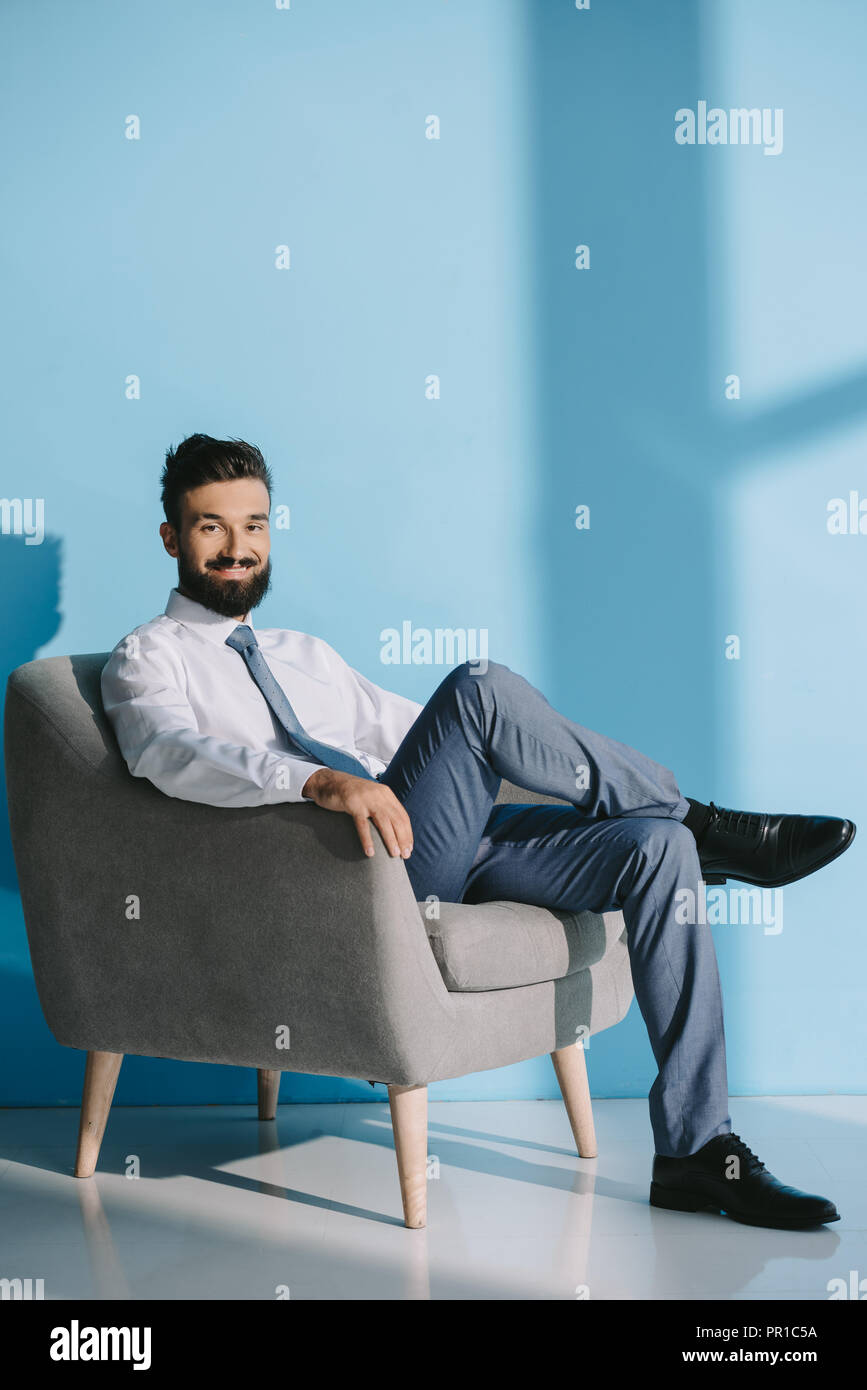 smiling businessman in formal wear sitting in grey armchair, on blue ...