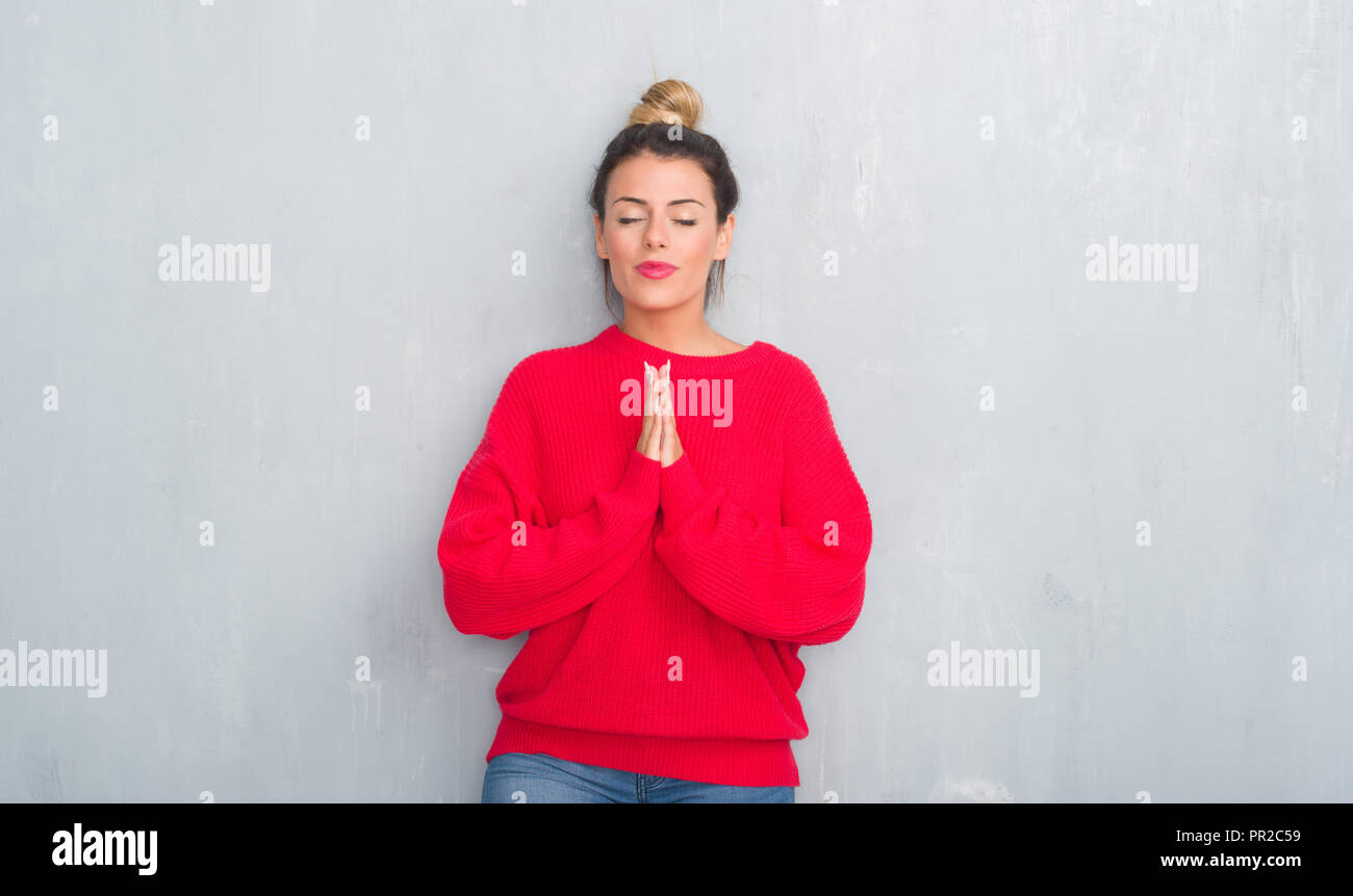 Young adult woman over grey grunge wall wearing winter outfit praying ...
