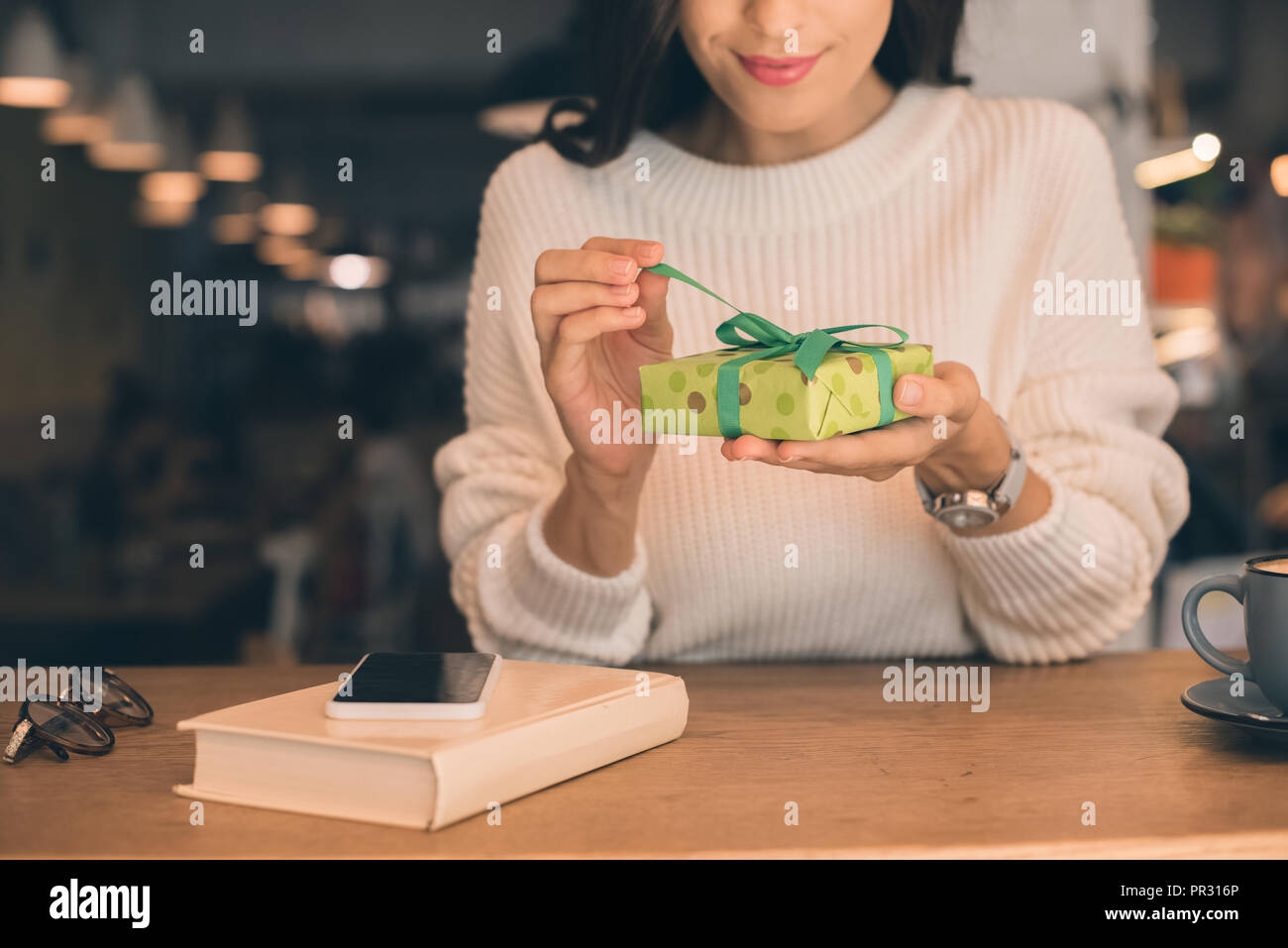 partial view of young woman untying gift box at table with smartphone ...
