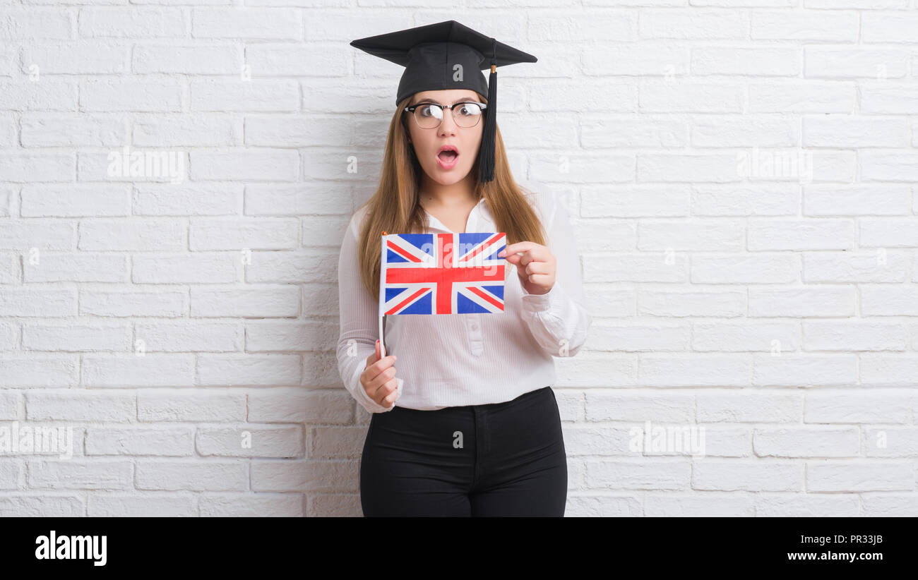 Young adult woman over white brick wall wearing graduate cap holding ...