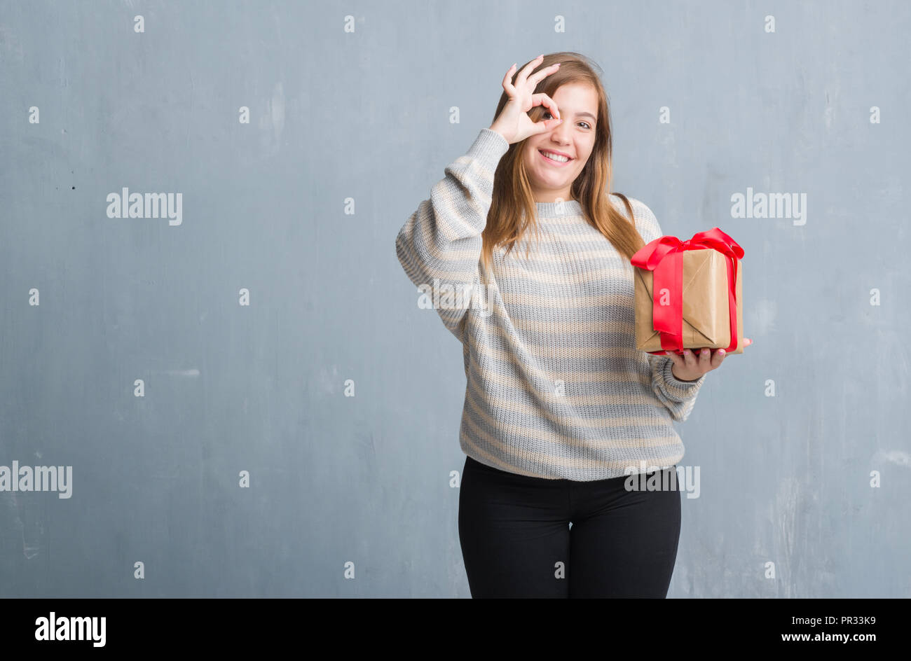 Young adult woman over grey grunge wall holding a present with happy ...