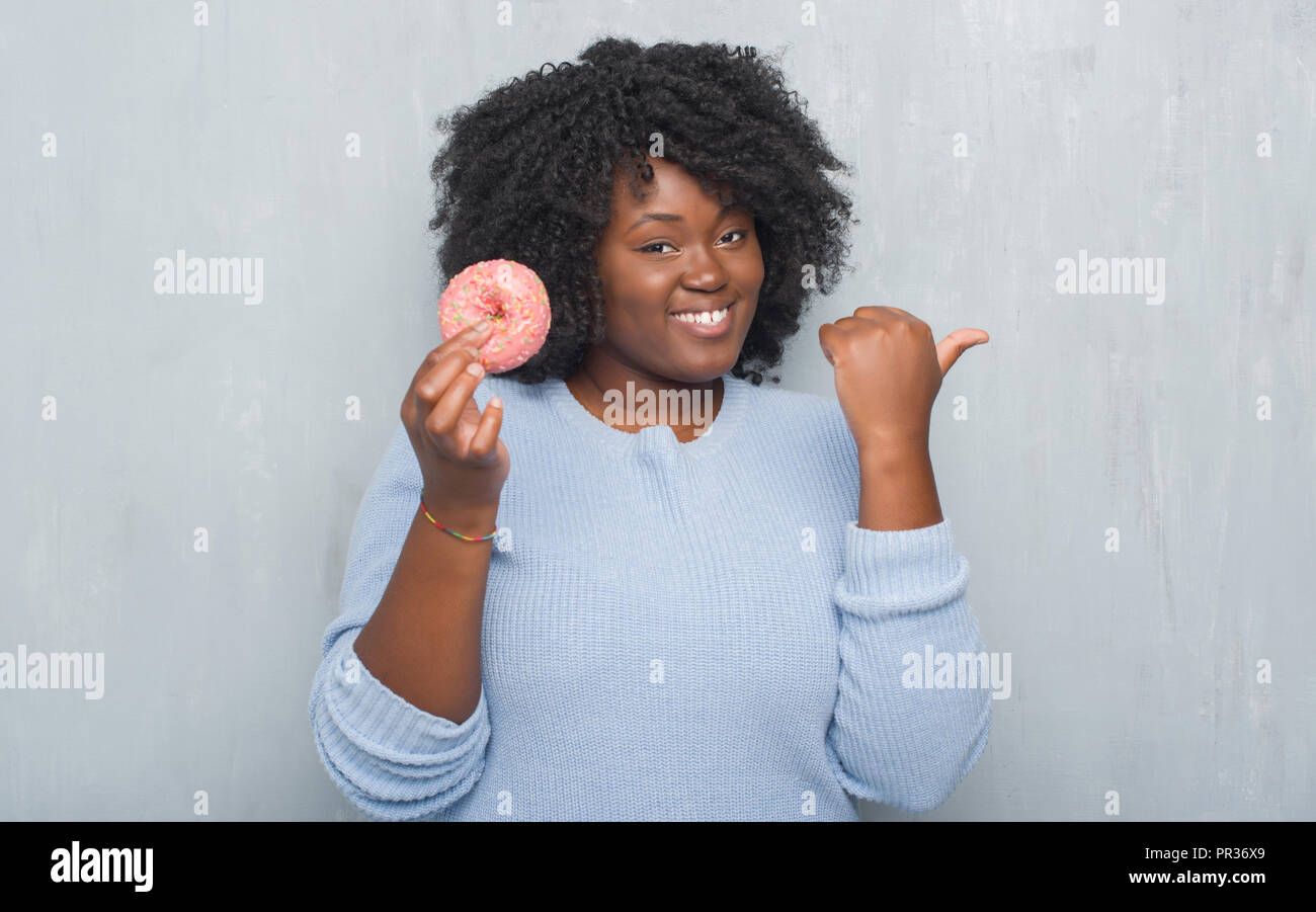 Young african american woman over grey grunge wall eating pink donut ...