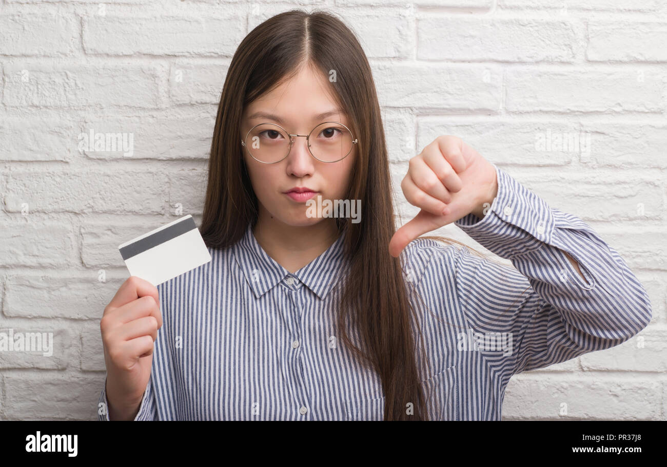 Young Chinese woman over brick wall holding credit card with angry face ...