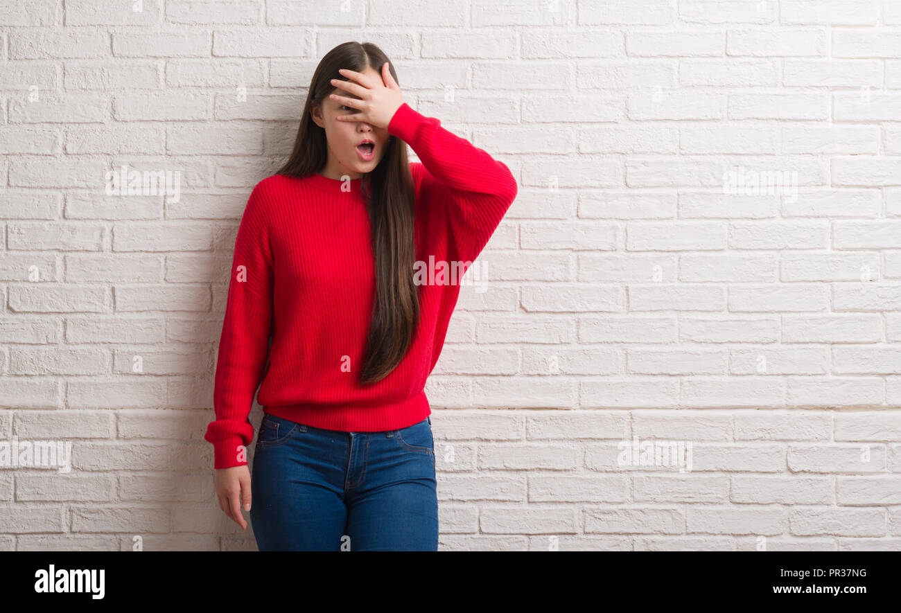 Young Chinese woman over brick wall peeking in shock covering face and ...