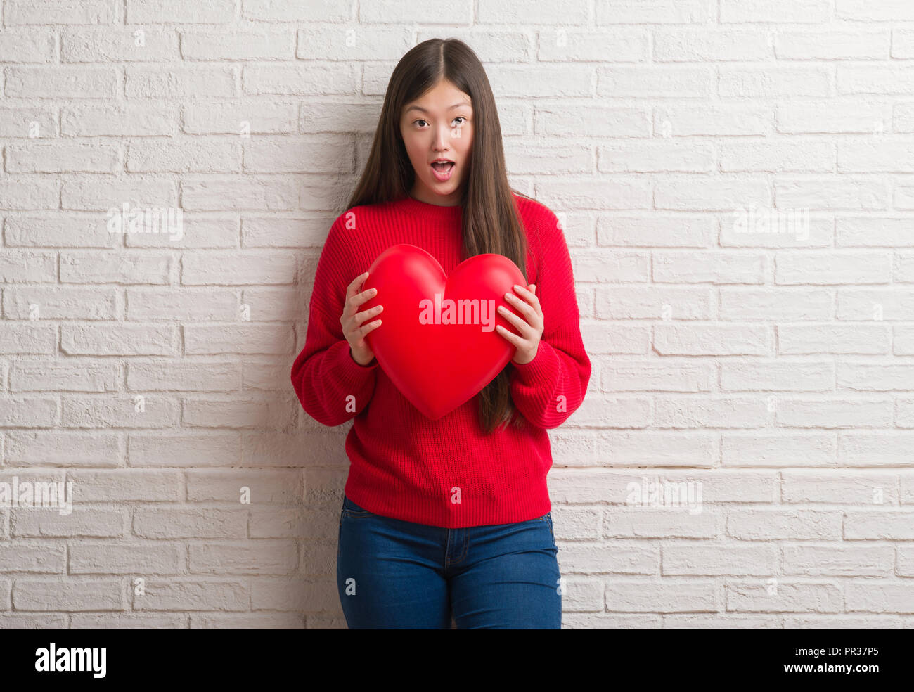 Young Chinese woman in love over brick wall holding red heart scared in ...