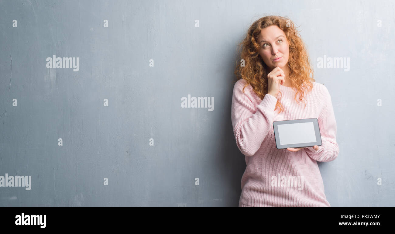 Young redhead woman over grey grunge wall using tablet serious face ...