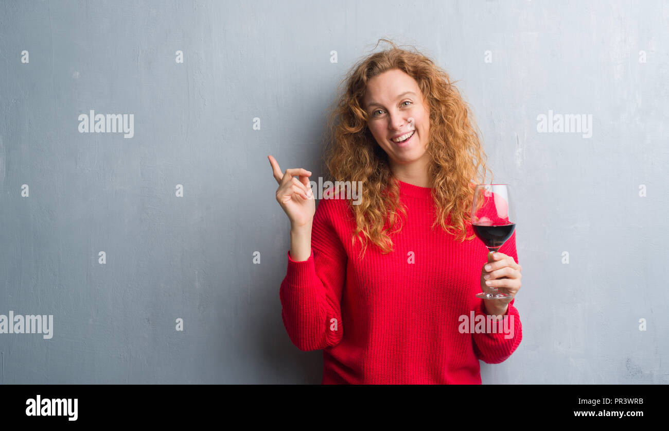 Young redhead woman over grey grunge wall drinking a glass of wine very ...