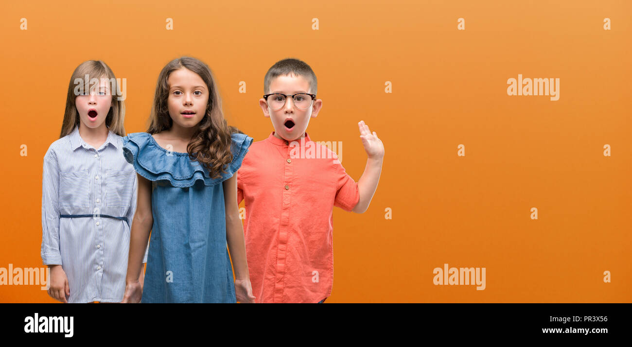 Group of boy and girls kids over orange background scared in shock with ...