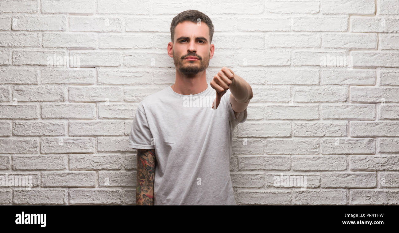 Young adult man standing over white brick wall with angry face ...
