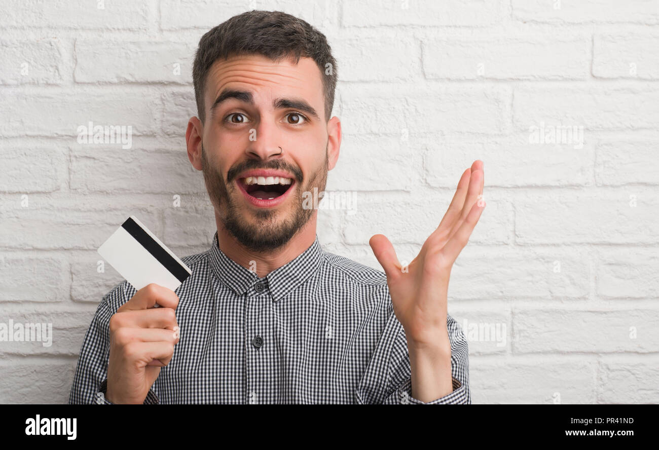 Young adult man over brick wall holding credit card very happy and ...