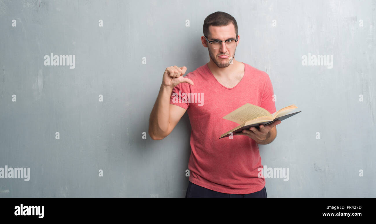Young caucasian man over grey grunge wall reading a book with angry ...
