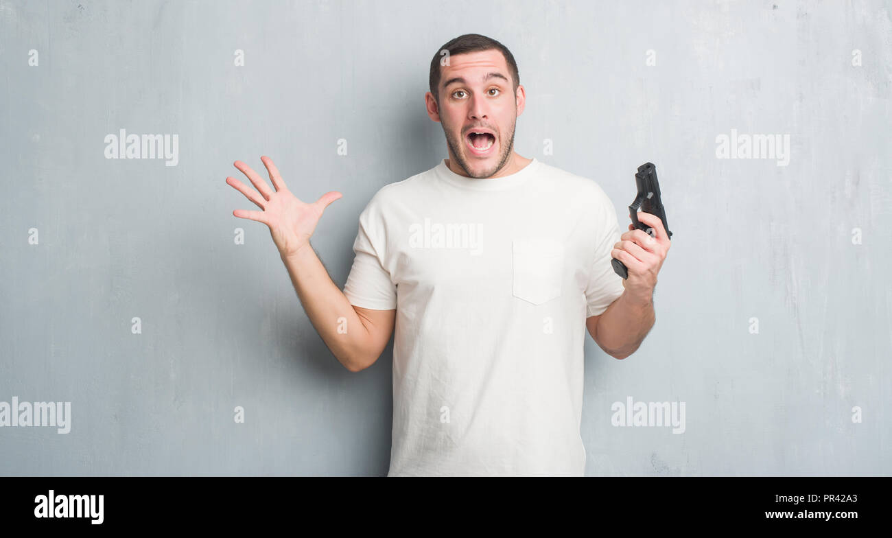 Young caucasian criminal man over grey grunge wall holding gun very ...