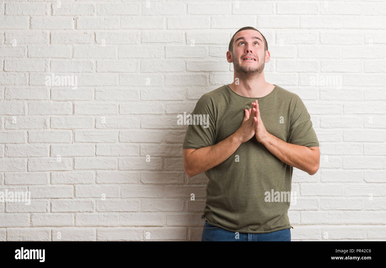 Young caucasian man standing over white brick wall begging and praying ...