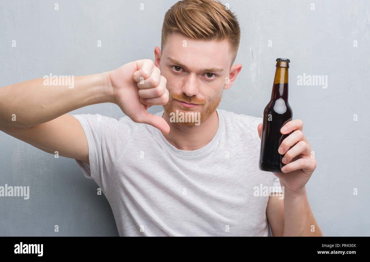 Young redhead man over grey grunge wall drinking beer bottle with angry ...