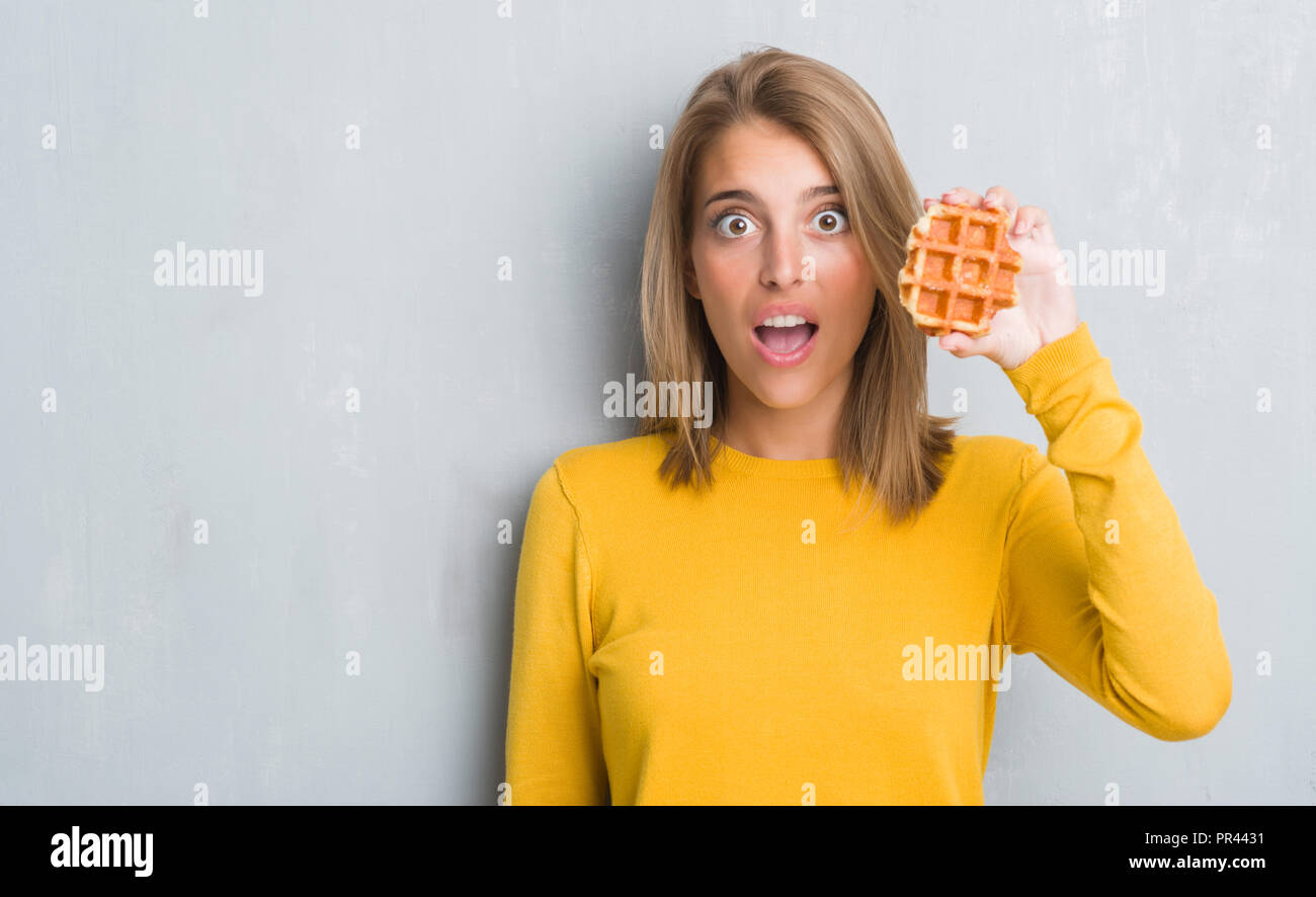 Beautiful young woman over grunge grey wall eating waffle scared in ...