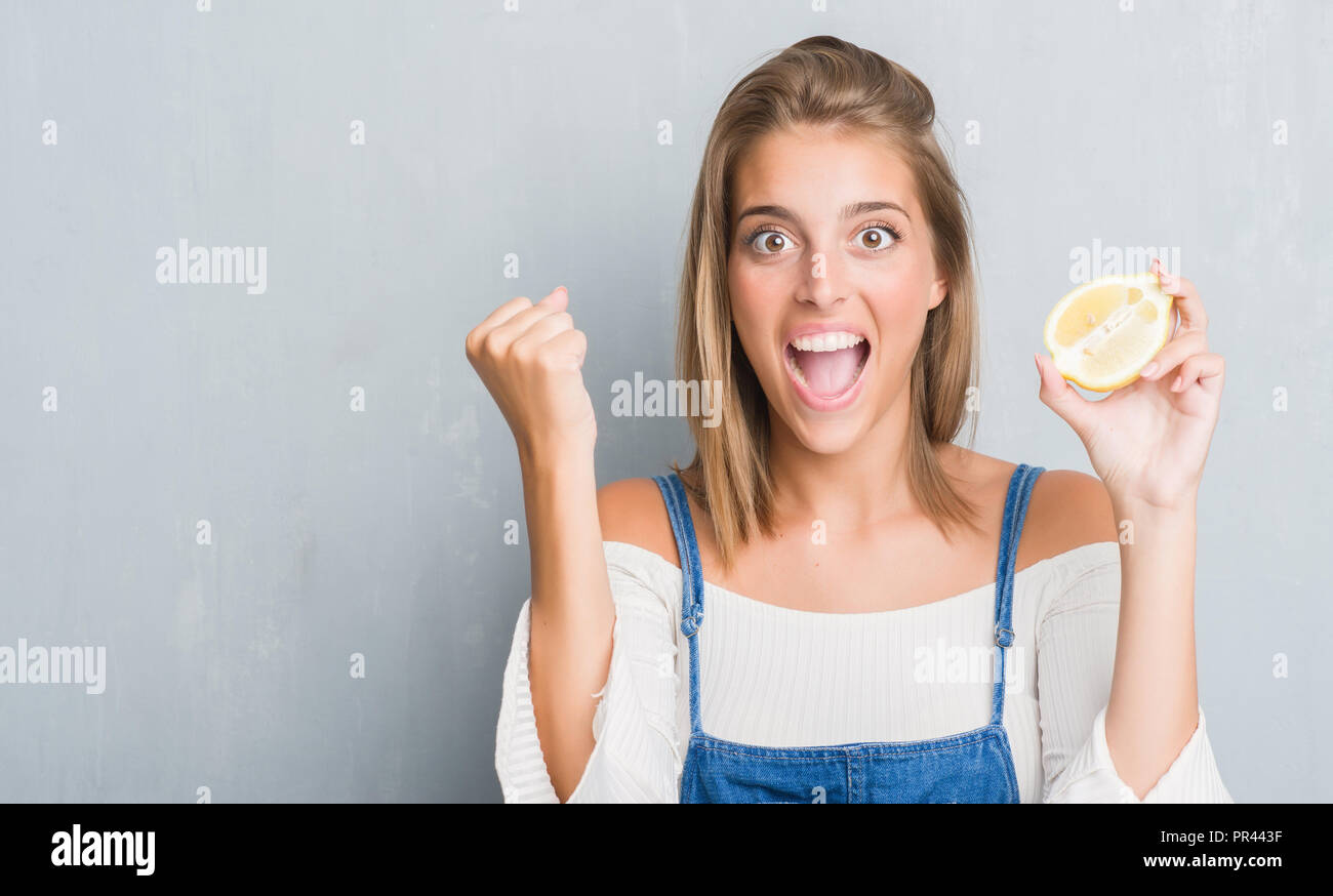 Beautiful young woman over grunge grey wall holding a lemon screaming ...