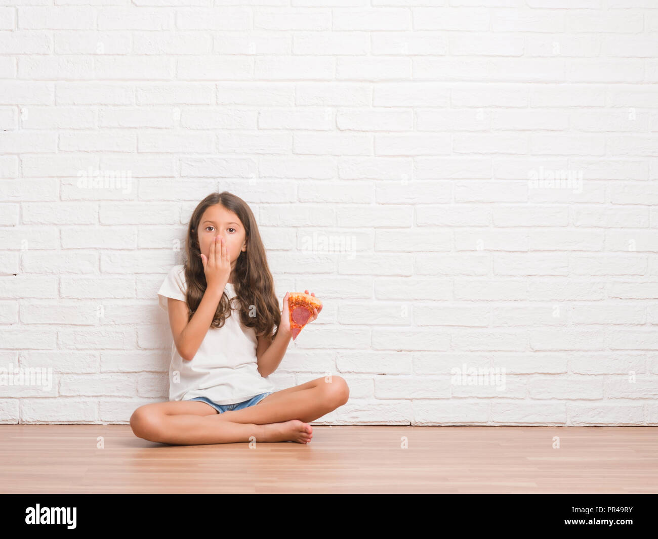 Young hispanic kid sitting on the floor over white brick wall eating ...