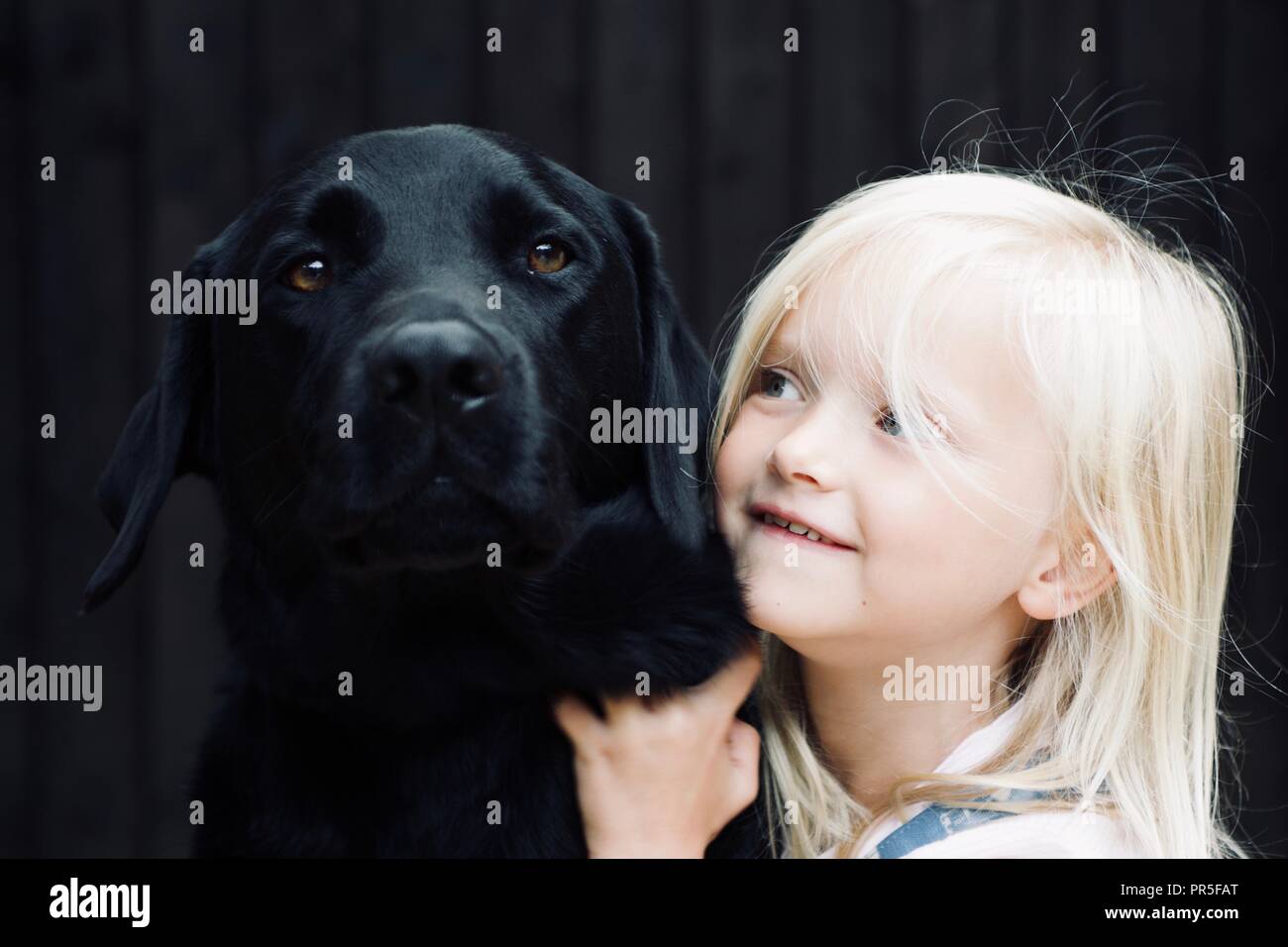 Young blonde girl lovingly cuddles her black Labrador dog, both facing ...