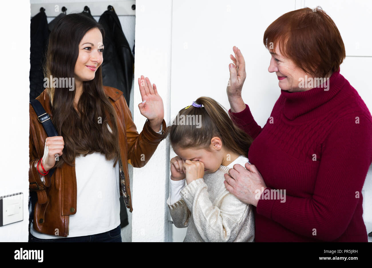 Smiling young mother leaving daughter in tears with grandma at home ...