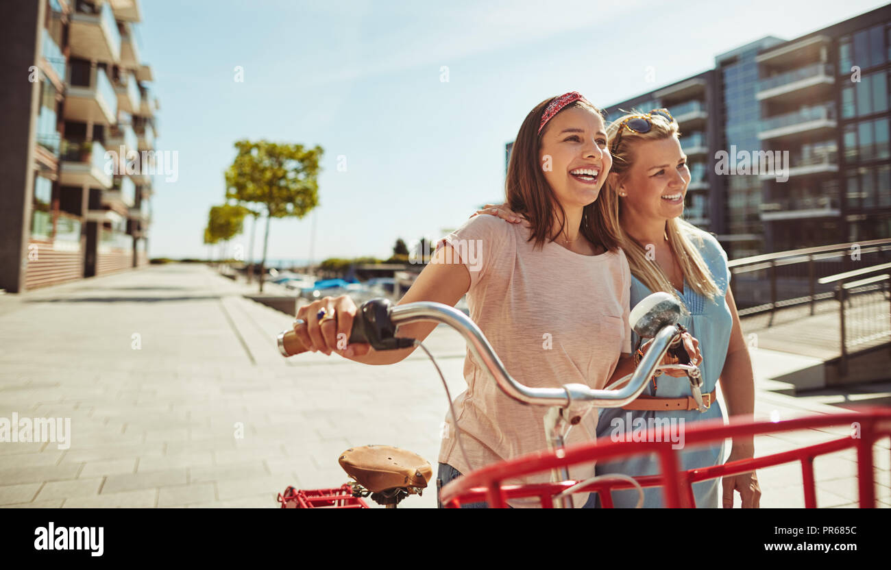 Two laughing young best friends walking with a bicycle through the city ...