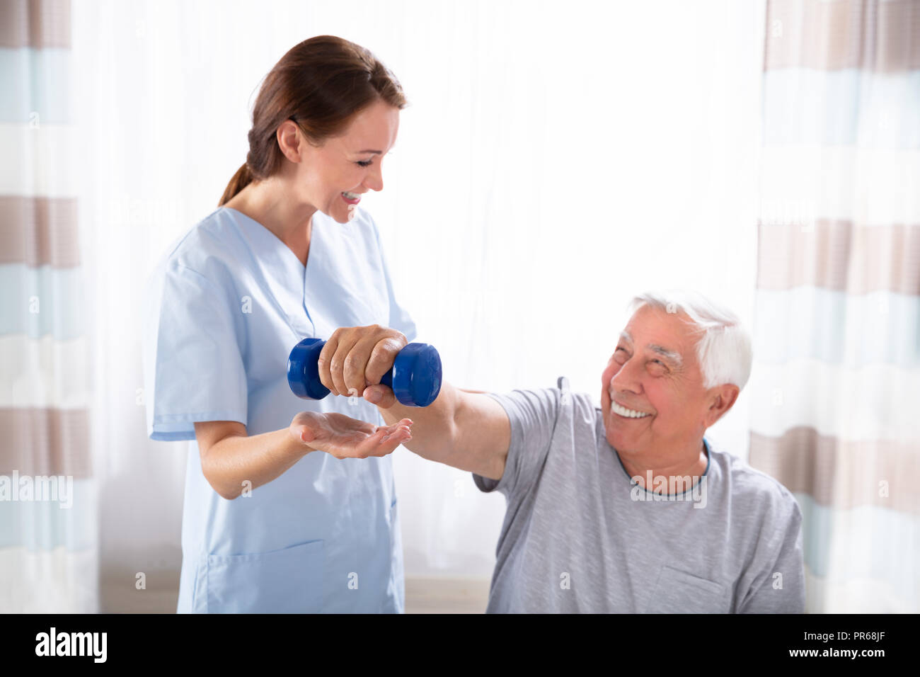 Young Female Nurse Helping Happy Senior Man With Dumbbell Exercise ...