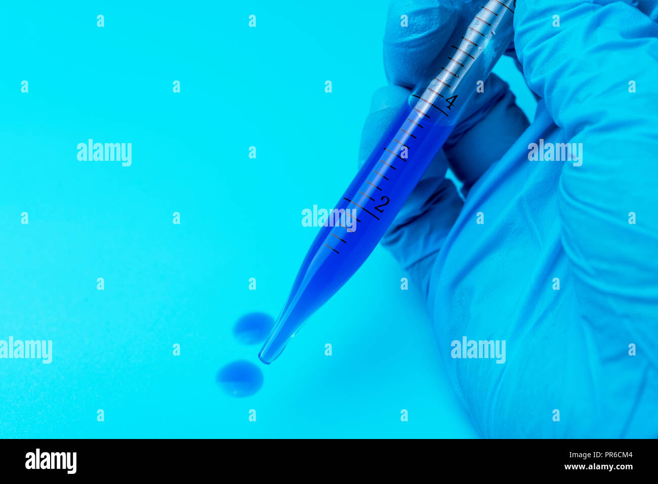 A gloved hand holds a lab pipette with blue liquid on a blue background ...