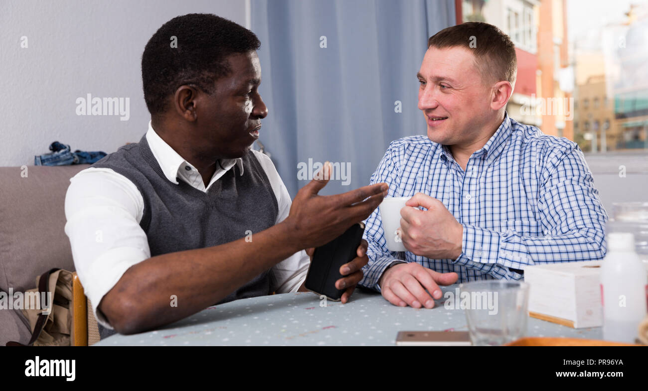 Two cheerful men enjoying conversation over cup of tea at home table ...