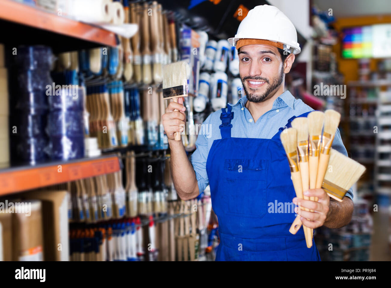 happy foreman in blue overalls choosing brushes in paint store Stock ...
