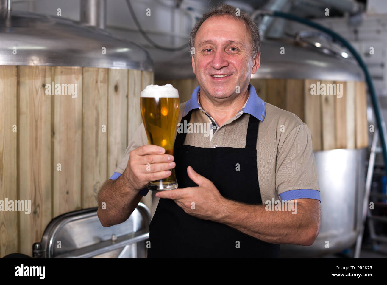 Brewer is standing with beer with foam in glass on his workplace indoor ...
