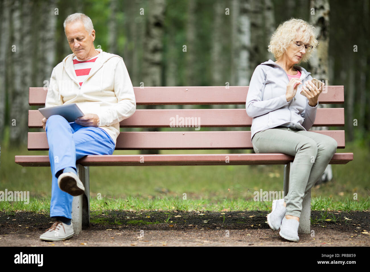 Senior man and woman sitting on opposite sides of bench and using ...