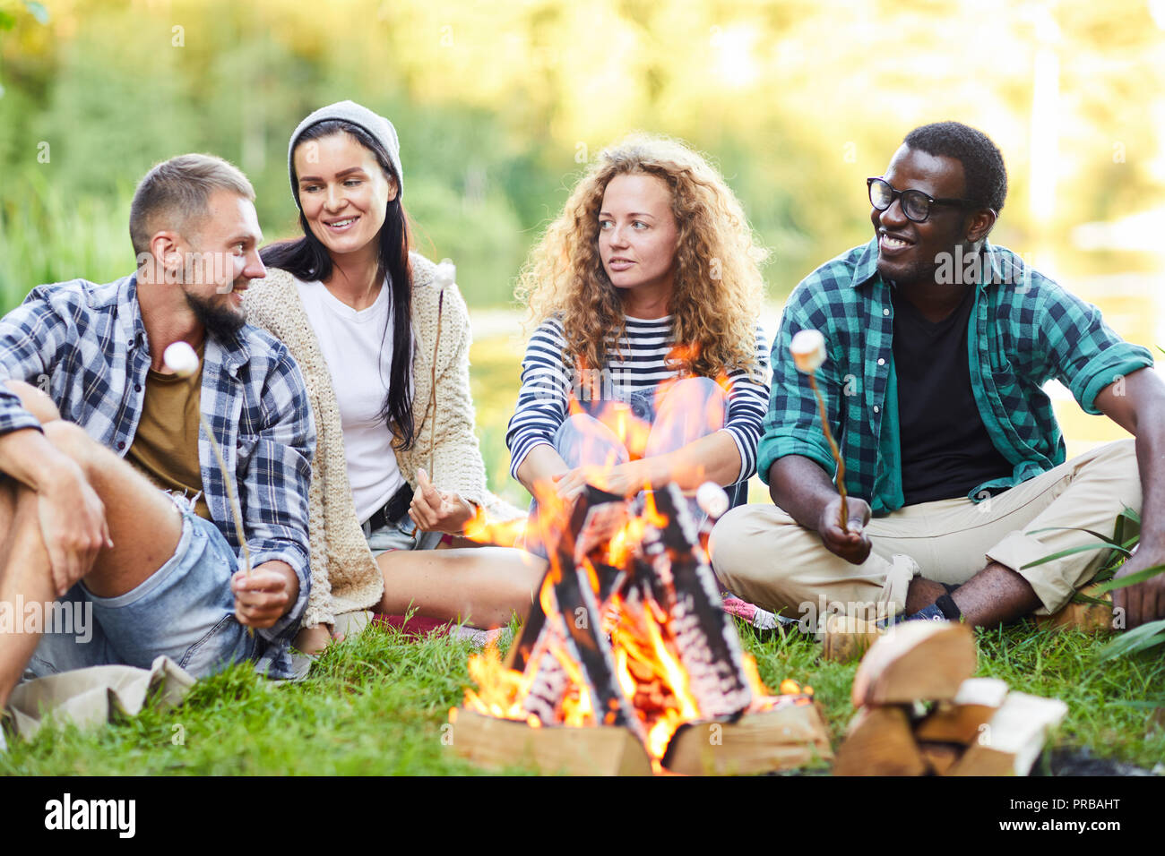 Group of young campers sitting by campfire and frying marshmellows ...