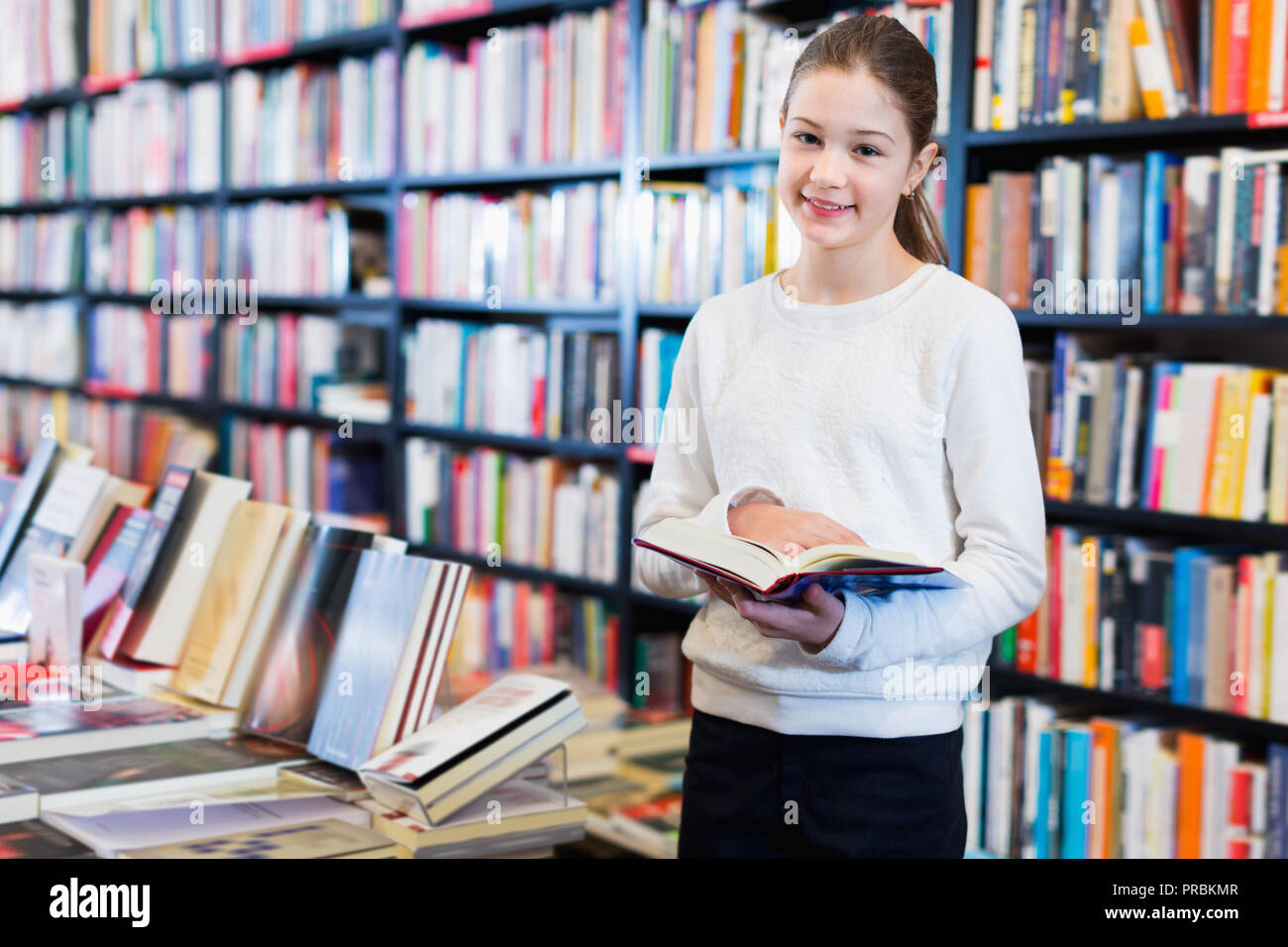 Intelligent cheerful preteen girl standing alone near bookcase in ...