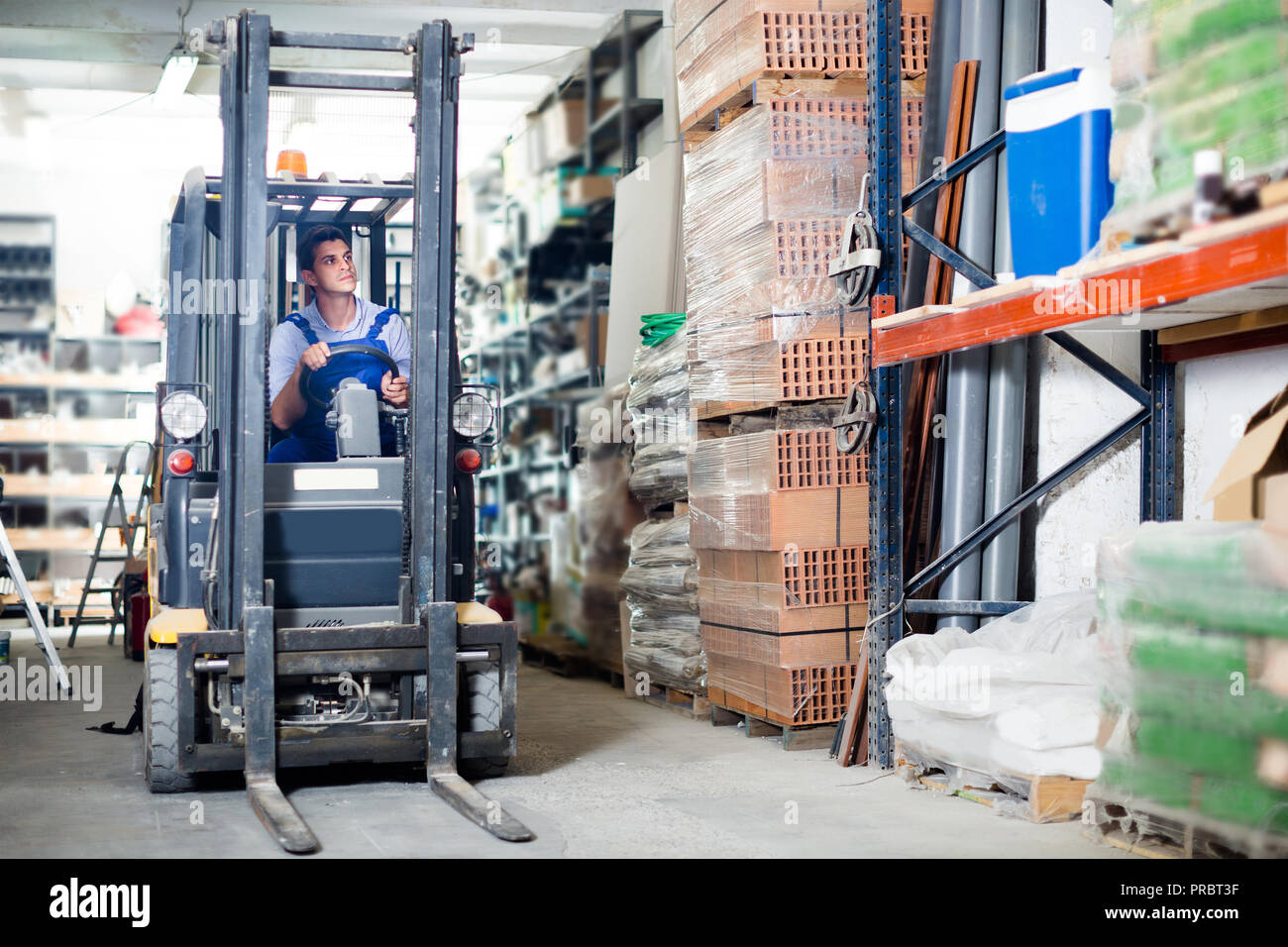 Shop assistant is driving loader between the rows in the building store ...