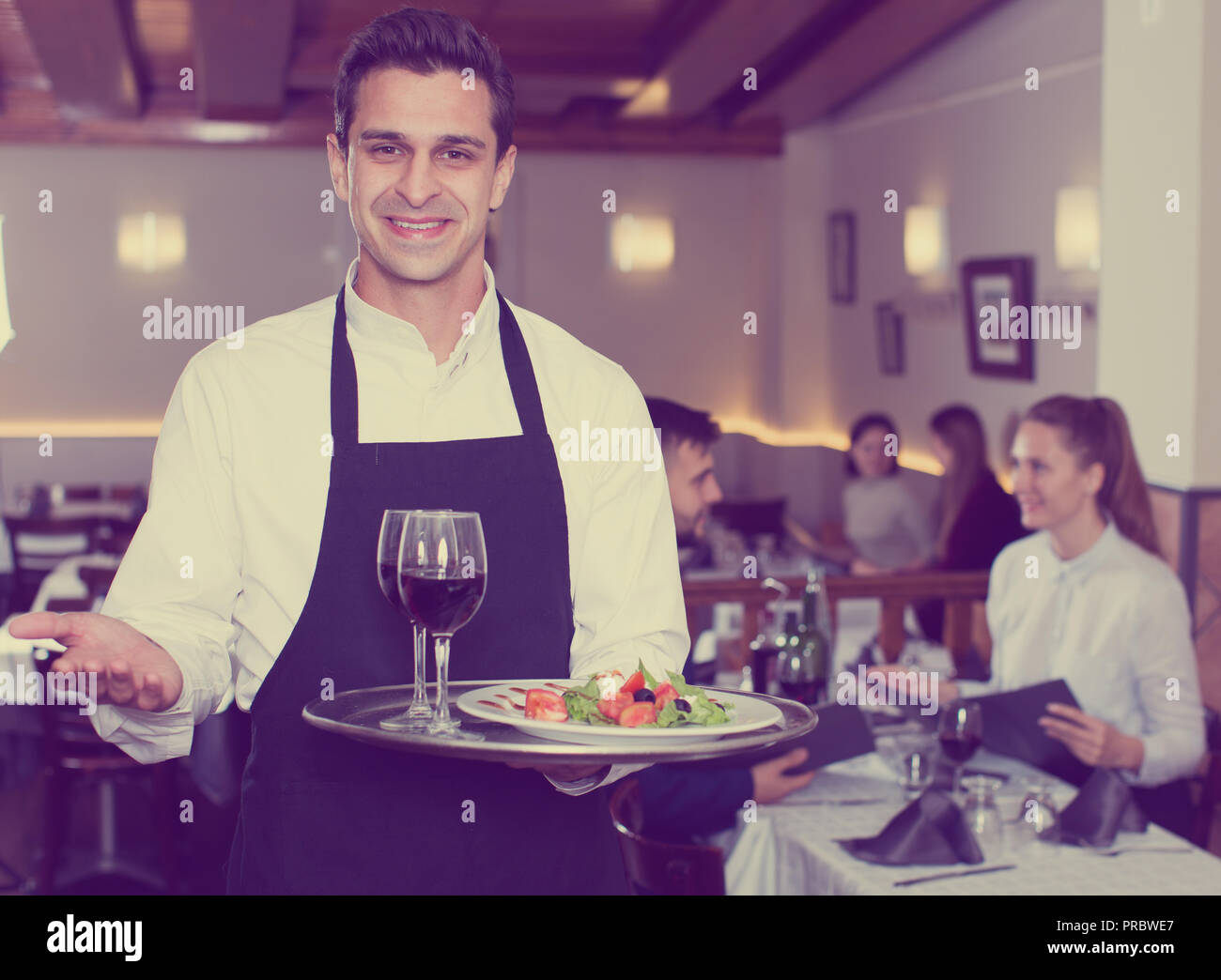 Polite waiter holding tray at restaurant with customers his behind ...