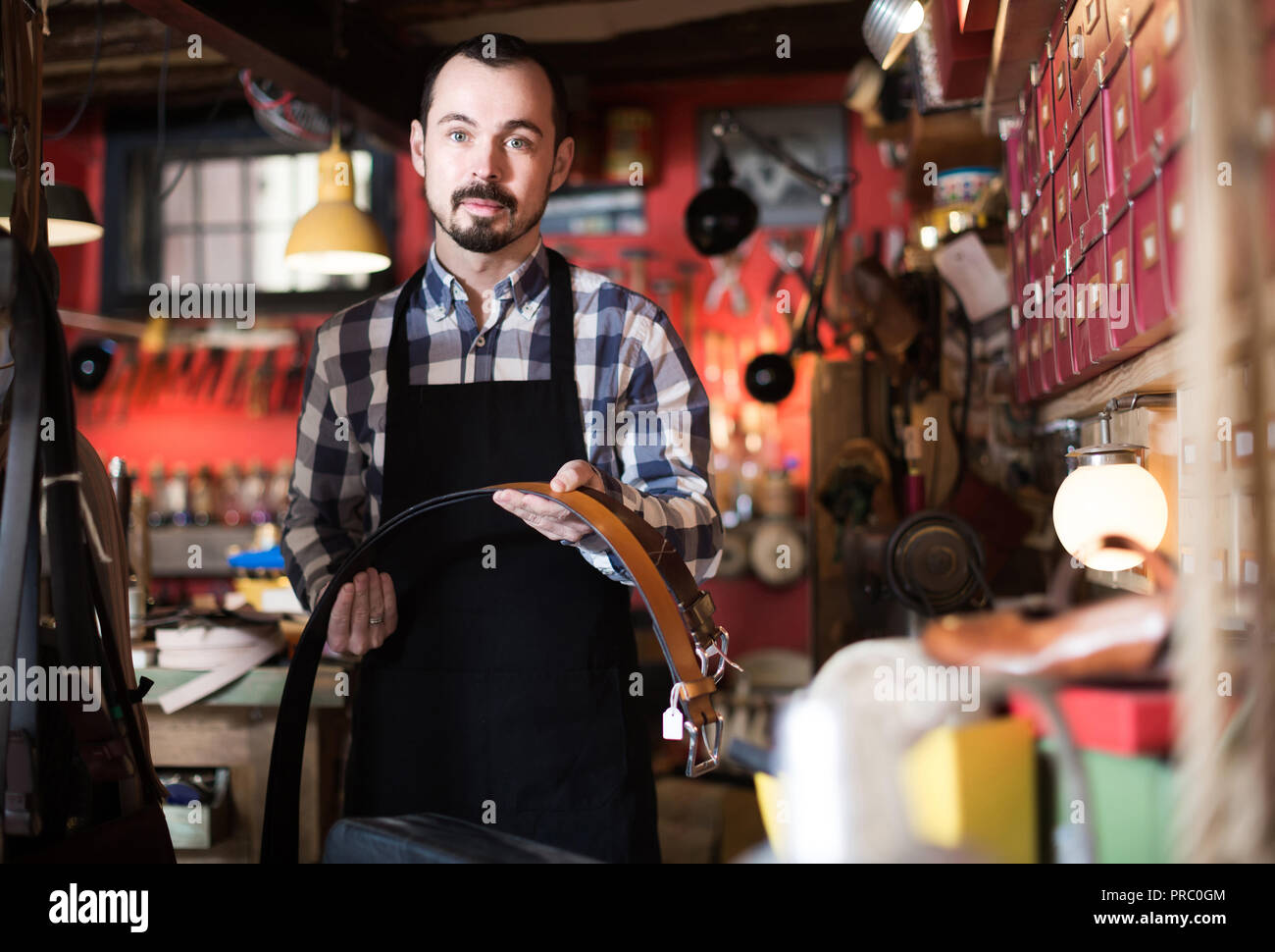 Young spanish man worker displaying various belts in leather workshop ...