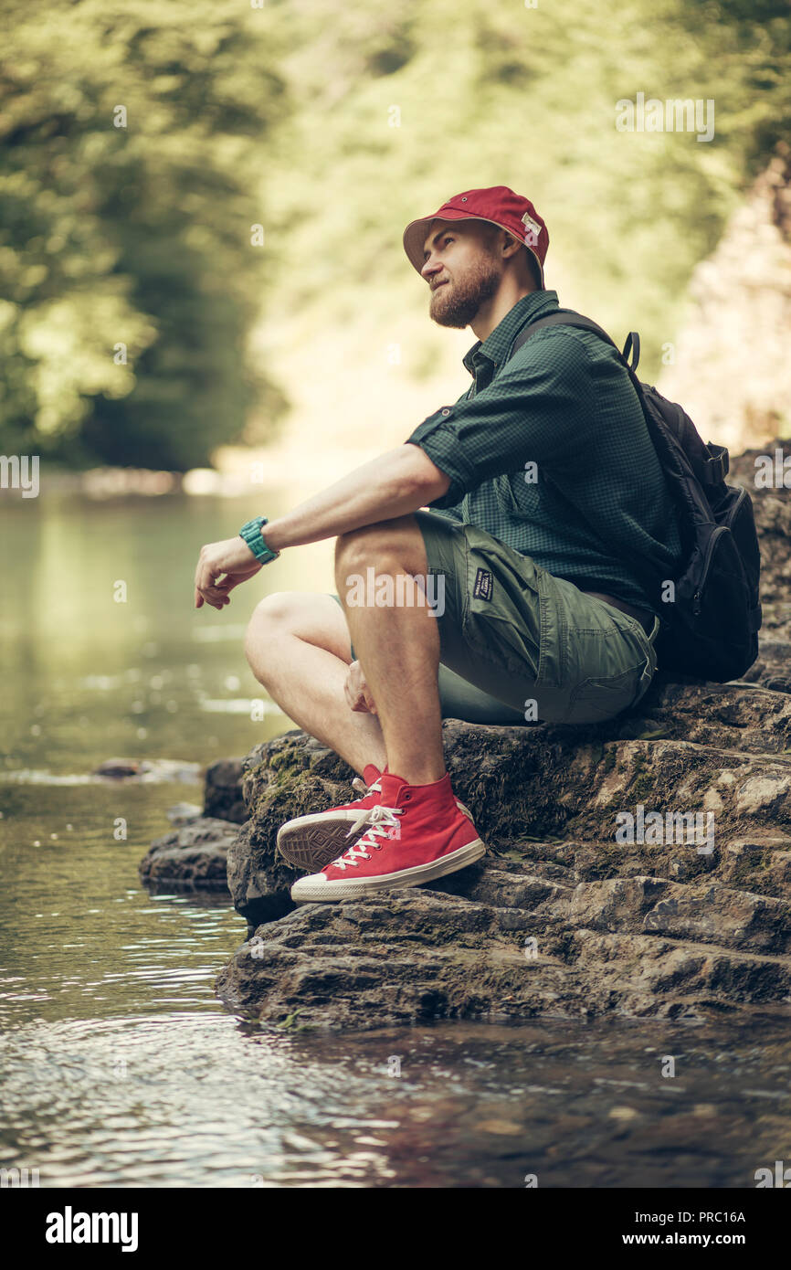 Tourist guy with backpack sitting near lake in a wild summer wood ...