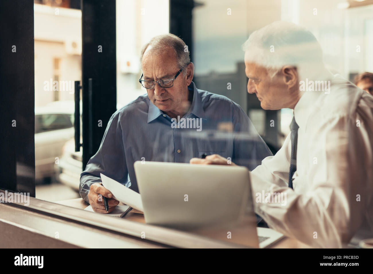 Shot through window glass of two mature businessman with laptop sitting ...