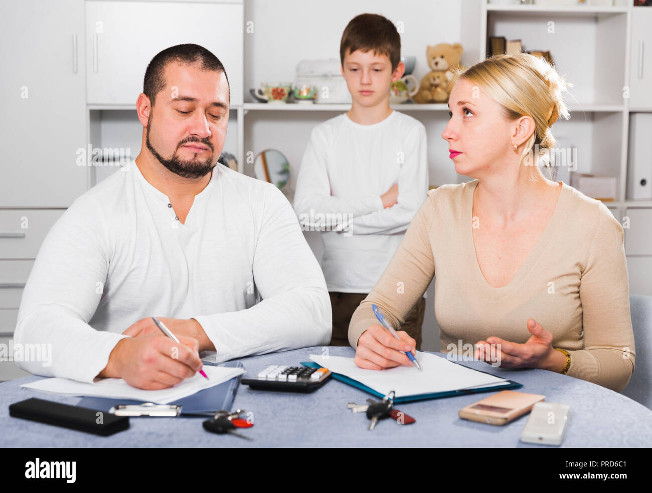Tense parents writing documents at home table with sad son behind Stock ...