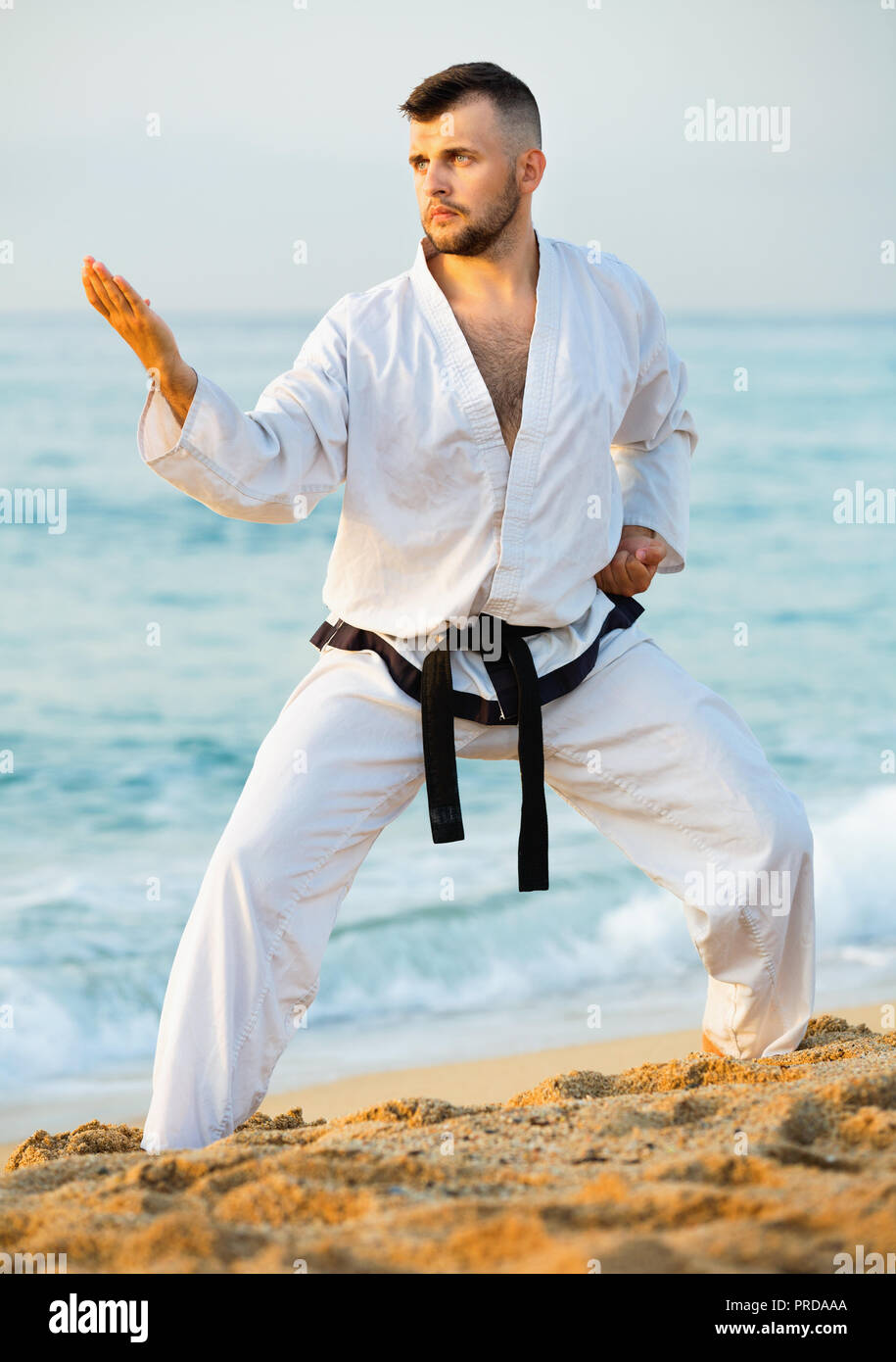 Glad positive man in uniform doing taekwondo exercises at sunset sea ...
