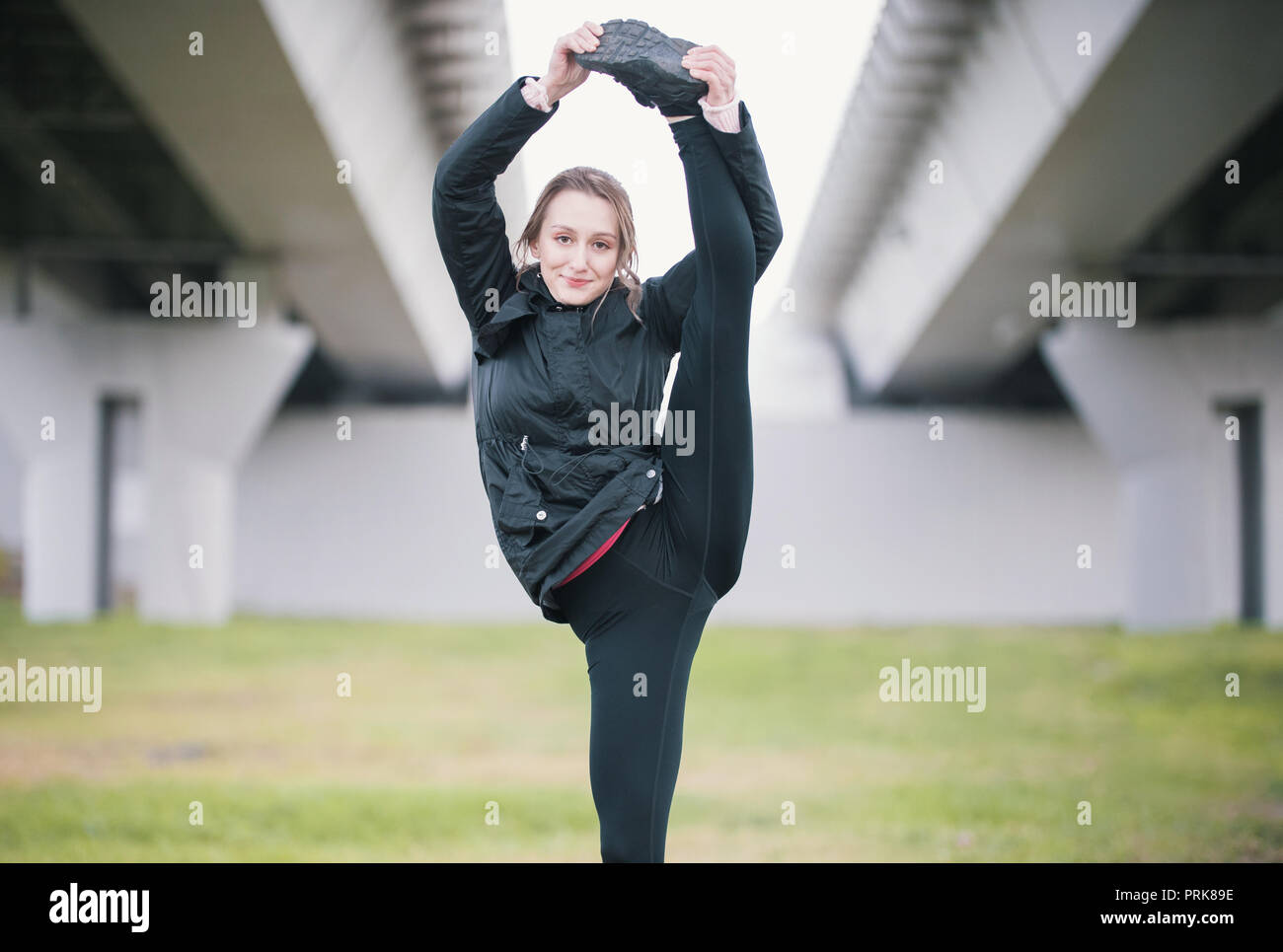 Young acrobat girl in black jacket performs stretching standing on one ...