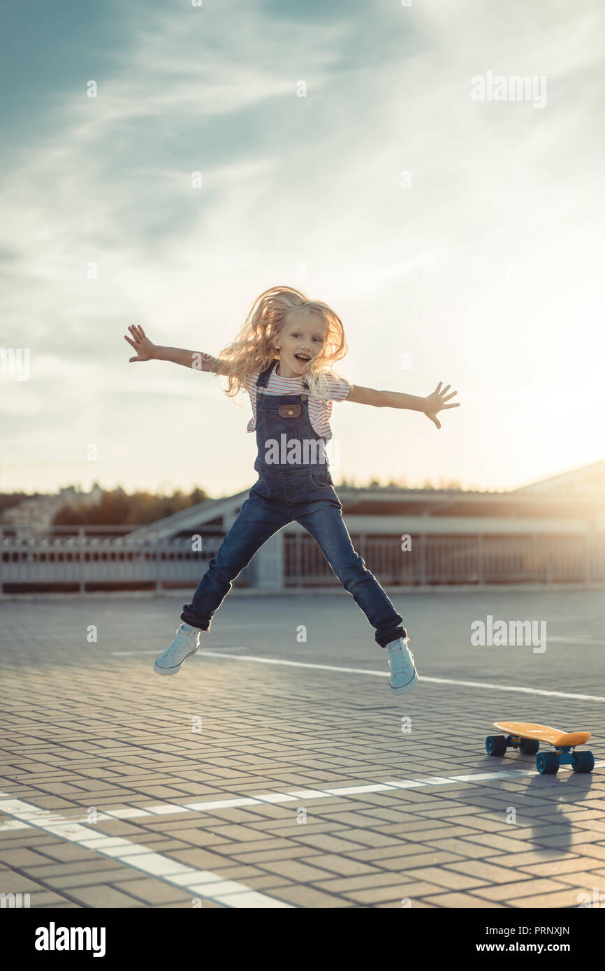 adorable little child jumping with wide arms near skateboard at parking ...